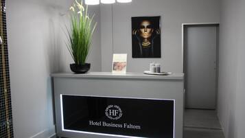 Bancada de recepção de hotel com uma planta em vaso, uma obra de arte emoldurada e uma placa com a inscrição 'Hotel Business Faltom'.