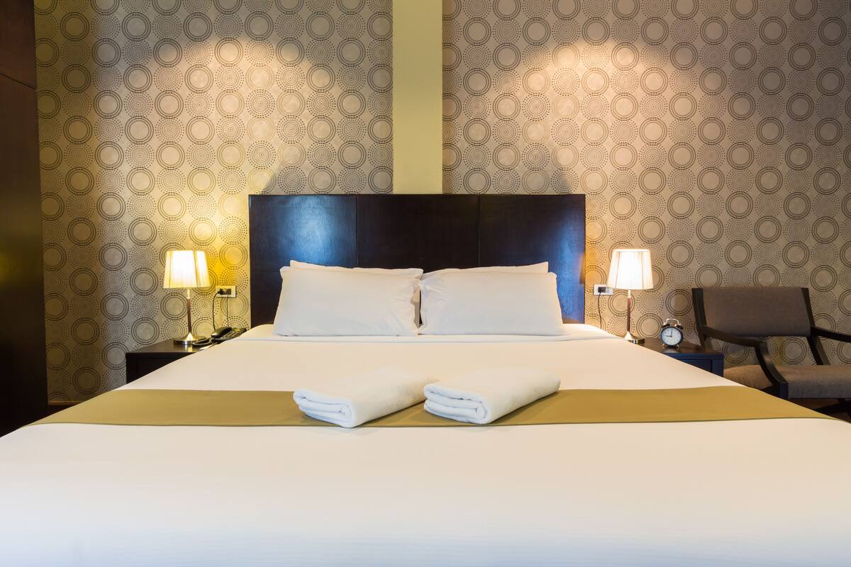 Uma cama bem arrumada com duas almofadas, duas toalhas dobradas e duas luminárias de cabeceira.