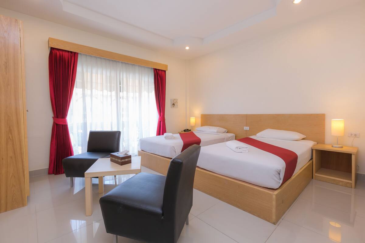 Quarto de hotel com duas camas, uma escrivaninha, uma cadeira e uma janela com cortinas vermelhas.