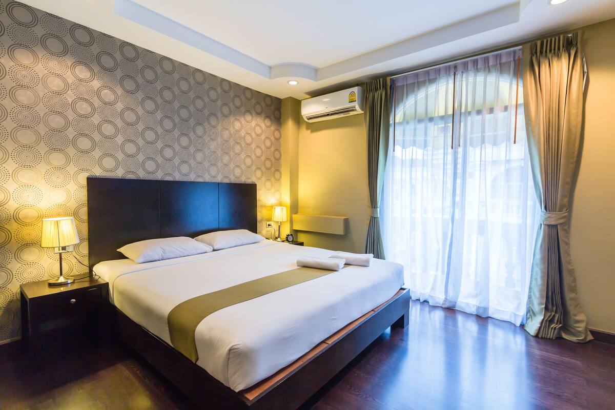 Quarto de hotel com uma cama grande, mesa de cabeceira com abajur, aparelho de ar condicionado e janela com cortinas.