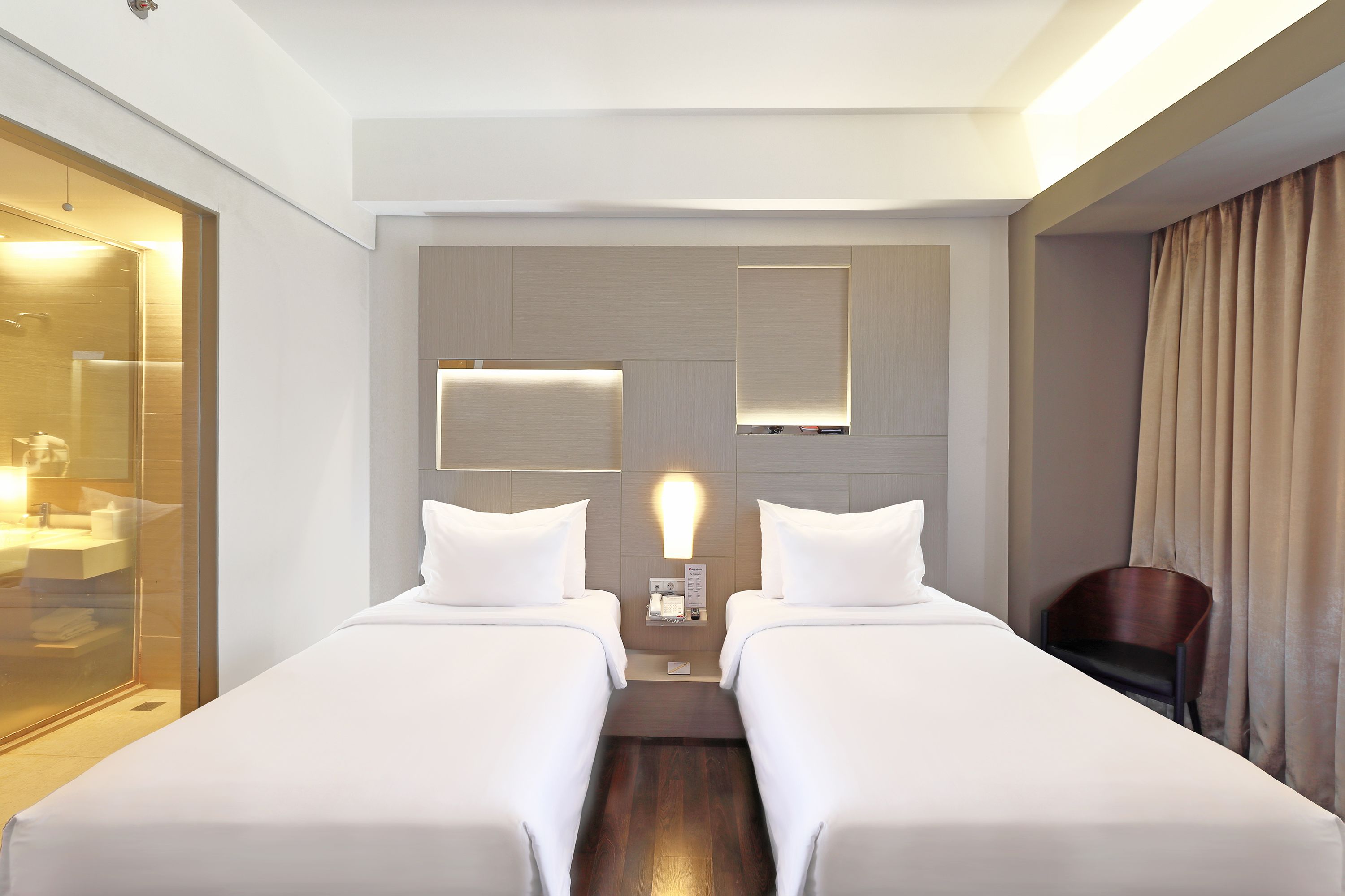 Quarto de hotel com duas camas de solteiro, mesa de cabeceira com abajur, espelho e uma cadeira.