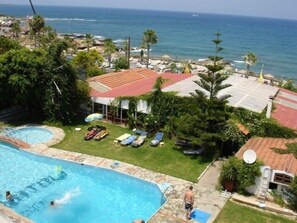 Piscina com vista para o oceano, rodeada por palmeiras e um edifício com telhado vermelho.