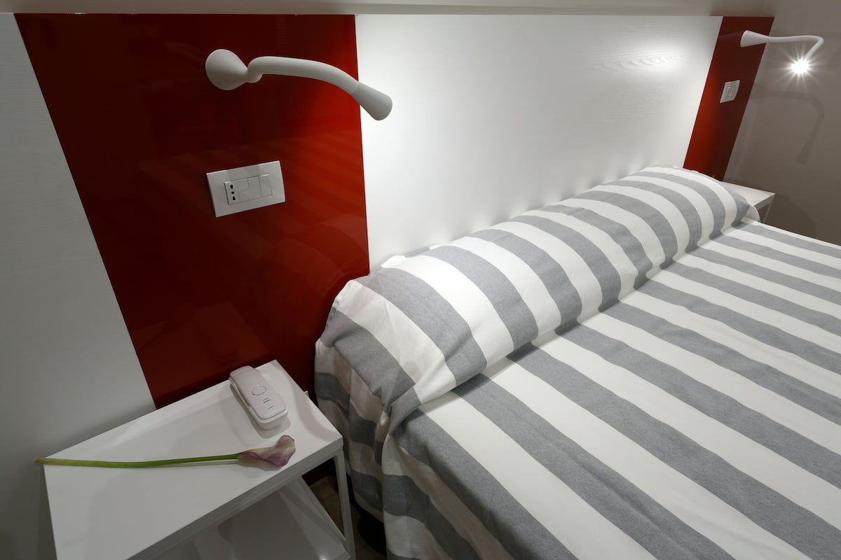 Quarto de hotel com cama listrada, parede de destaque vermelha, mesa de cabeceira branca com telefone e luminária branca.