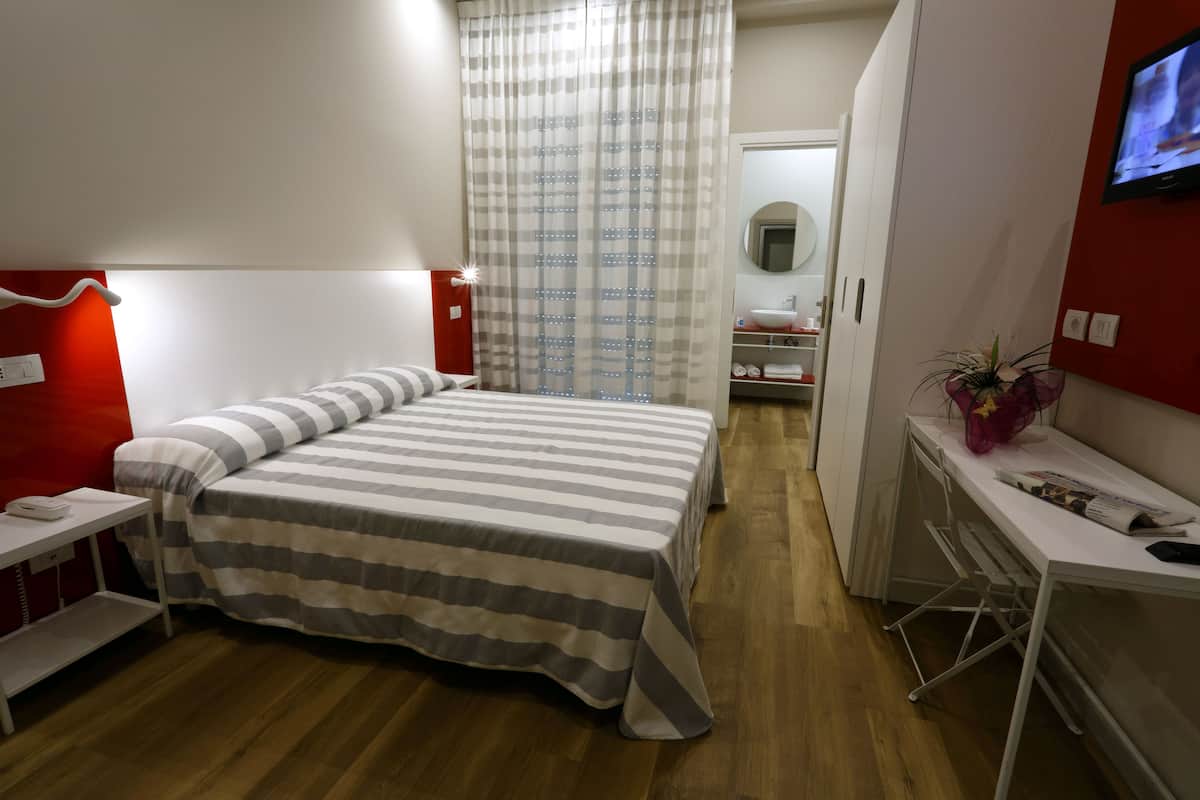 Quarto de hotel com cama, mesa de cabeceira, escrivaninha e televisão na parede.