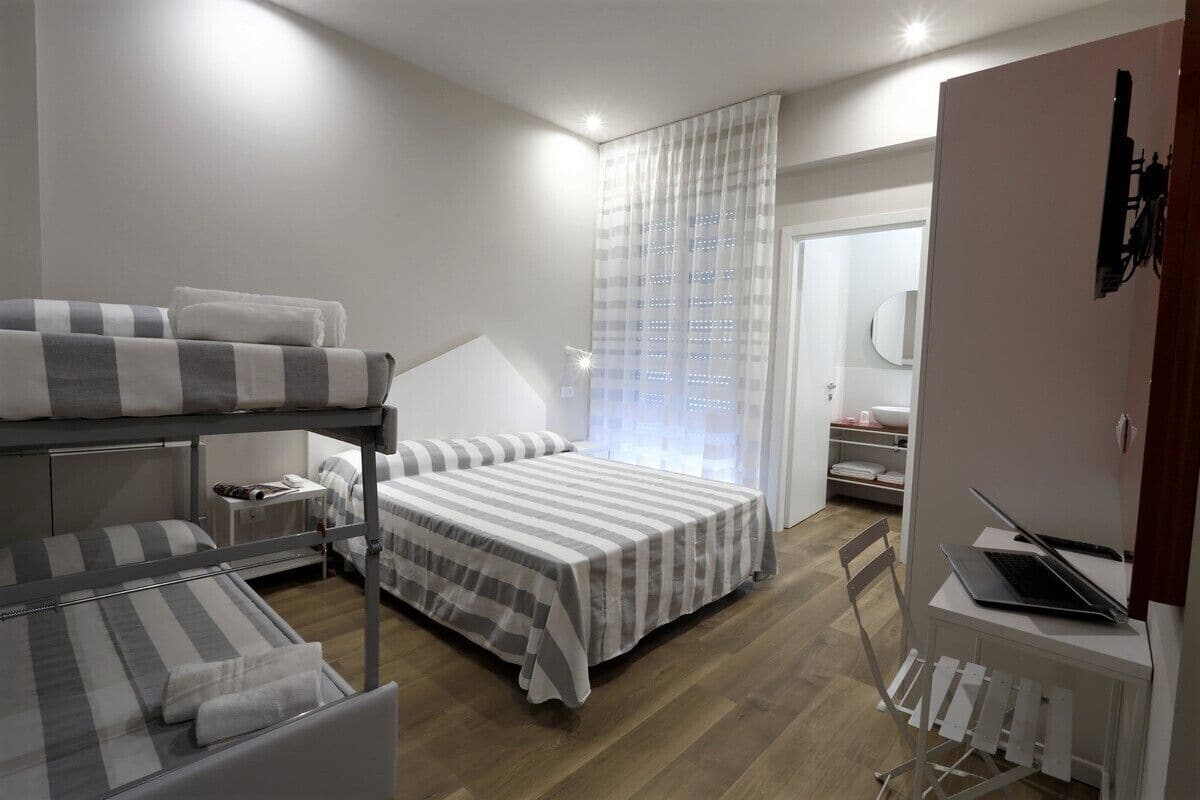 Quarto com uma cama beliche, uma cama de solteiro, uma escrivaninha com um laptop e um banheiro visível através de uma porta.