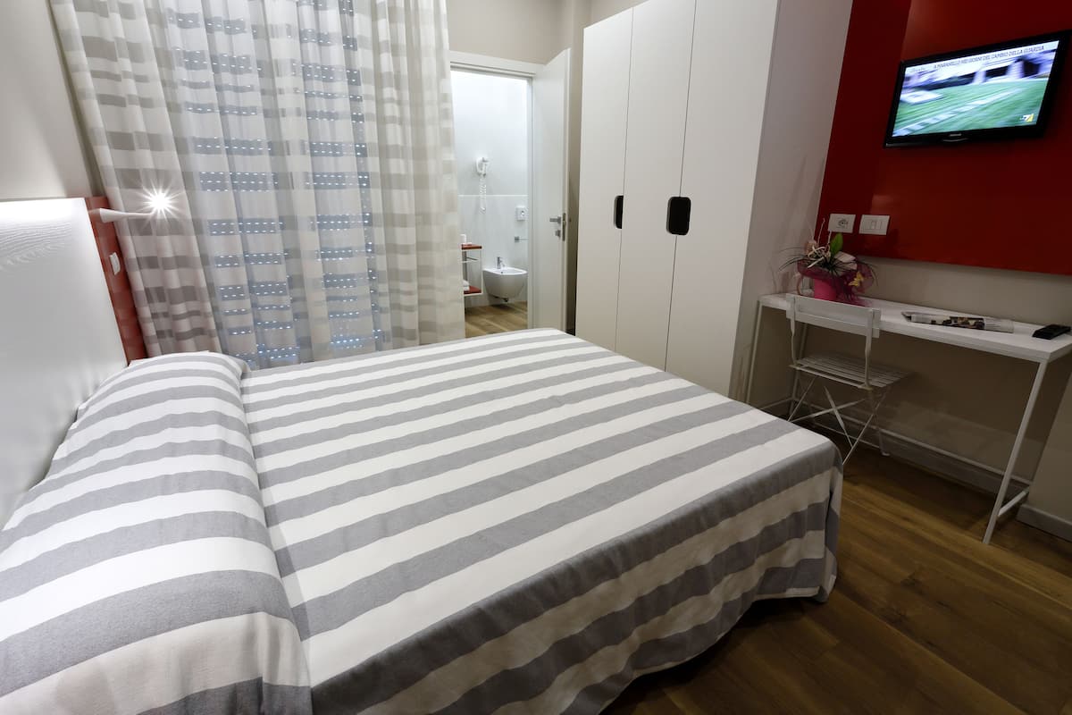 Quarto de hotel com uma cama, televisão na parede, escrivaninha e banheiro visível por uma porta aberta.