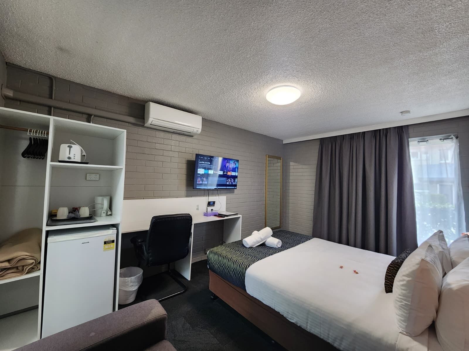 Quarto de hotel com cama, escrivaninha, cadeira, televisão e armário.