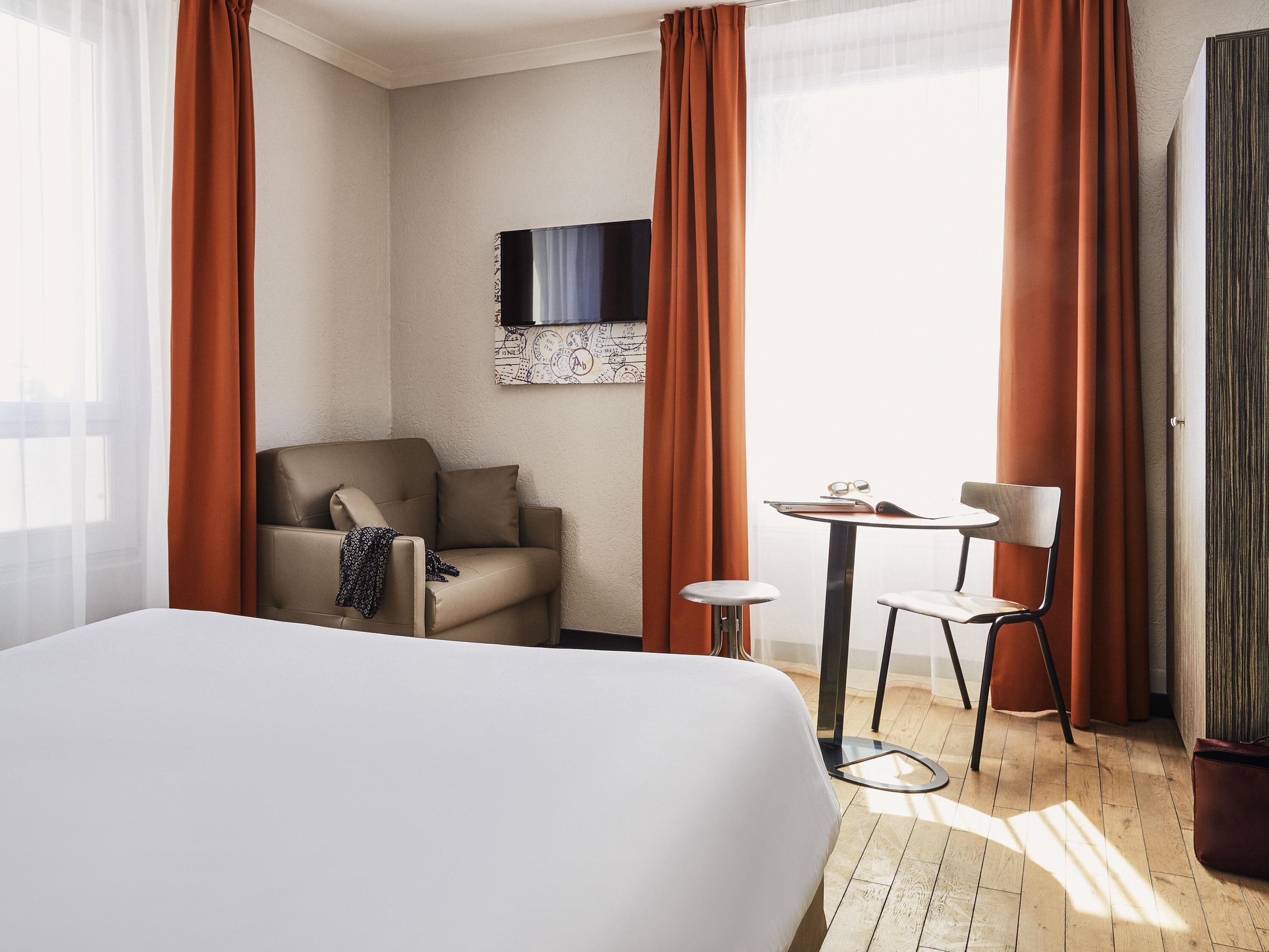 Quarto de hotel com cama, sofá, uma mesinha com cadeira e televisão na parede.
