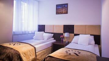 Quarto de hotel com duas camas, mesa de cabeceira, abajur, janela com cortinas translúcidas e um quadro na parede.