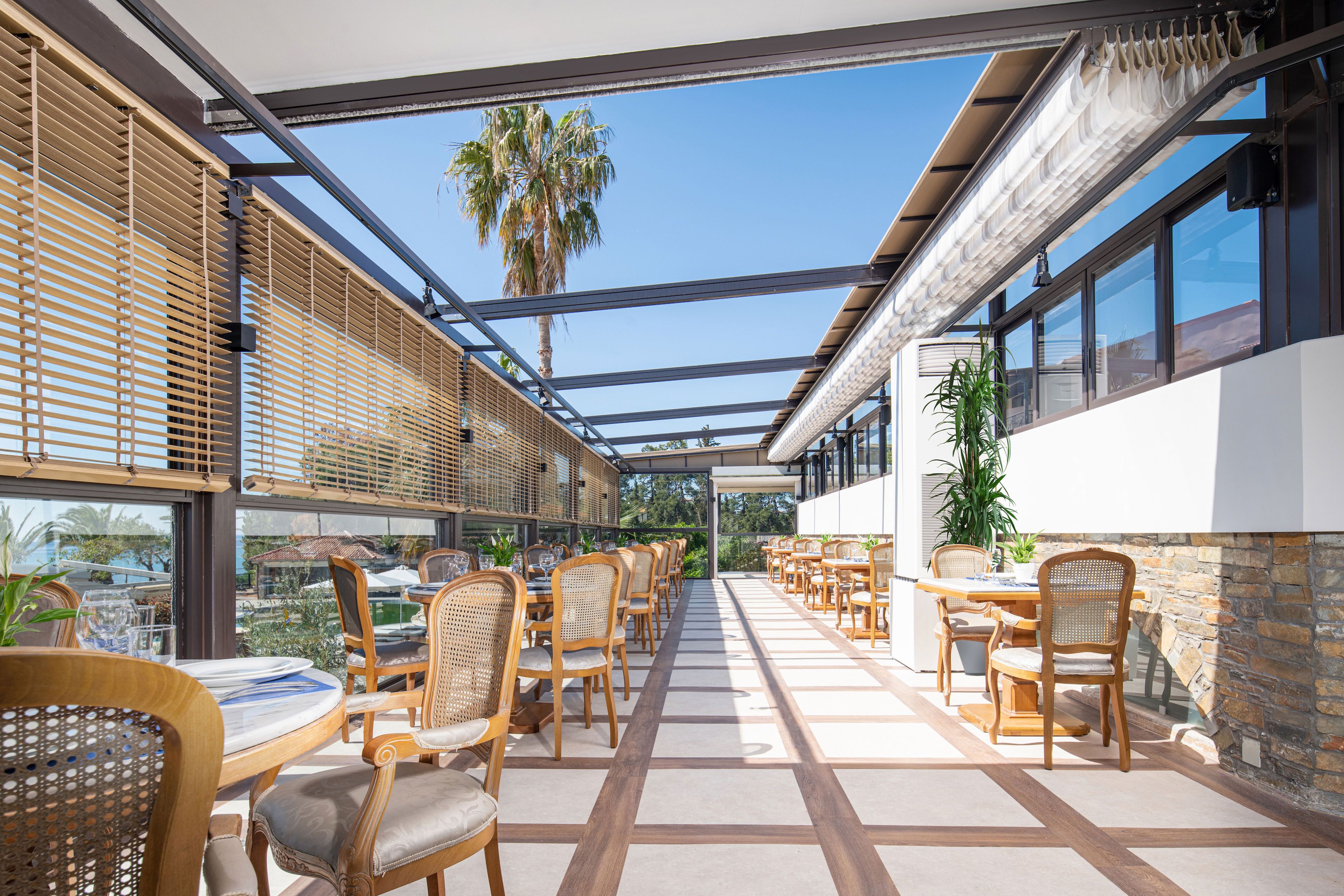 Uma área de refeições ao ar livre com mesas e cadeiras de madeira, vista desobstruída para palmeiras e um design arquitetônico moderno com grandes janelas e teto de vidro.