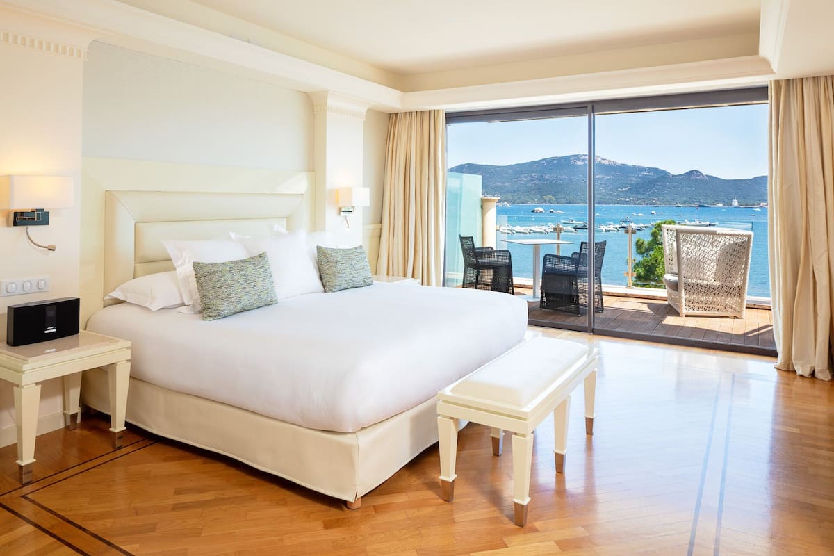 Um quarto espaçoso com uma cama grande, uma mesinha de cabeceira com um rádio e vista para o mar e barcos através de uma porta de vidro.