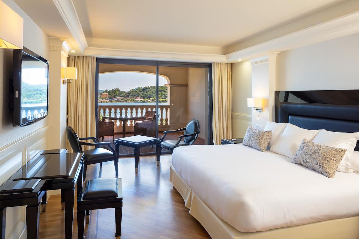 Quarto de hotel com uma cama grande, uma escrivaninha, duas cadeiras, televisão e varanda com vista panorâmica.