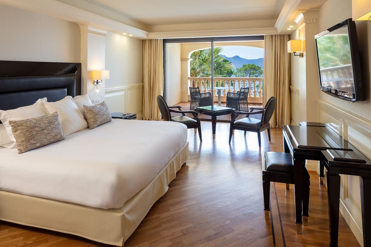 Quarto de hotel com uma cama grande, uma escrivaninha, uma cadeira e uma televisão. Possui uma varanda com vista para as montanhas.
