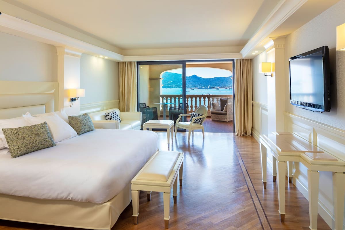Quarto de hotel com uma cama grande, televisão, varanda com vista para o mar e área de refeições.