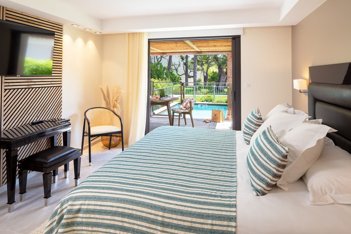 Quarto de hotel com uma cama grande, uma mesa com cadeira, uma televisão e vista para uma piscina e vegetação lá fora.