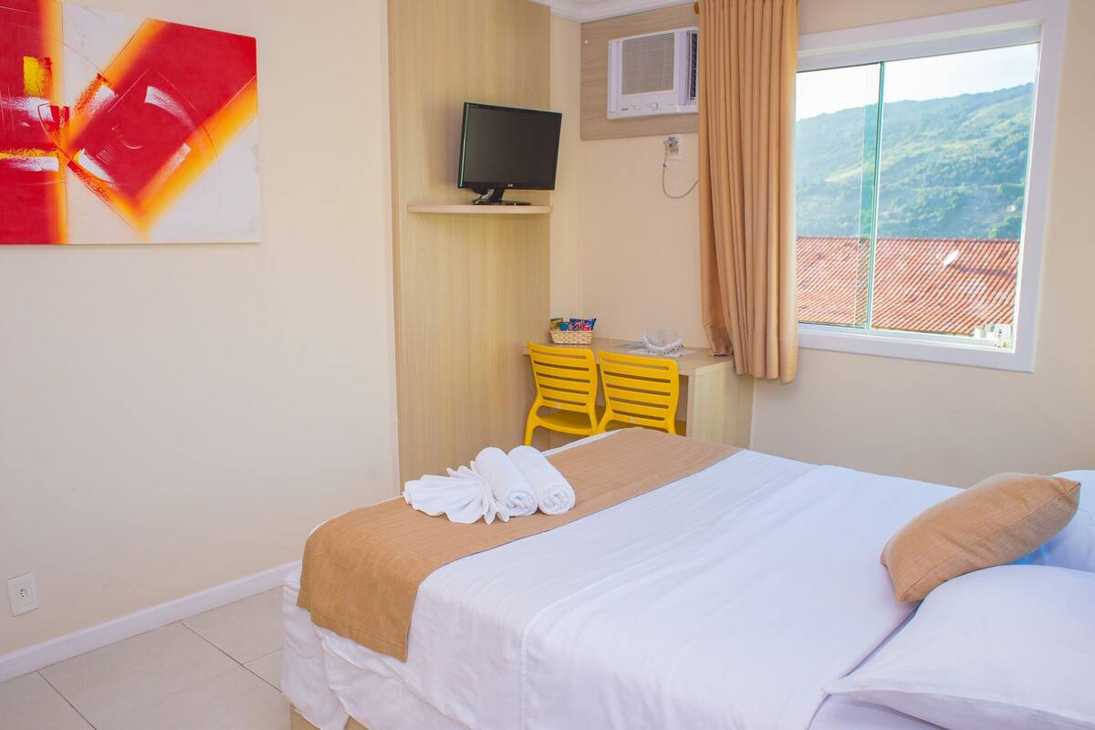 Quarto de hotel com uma cama, uma televisão presa na parede, uma mesa com cadeira e uma janela com vista para as colinas.