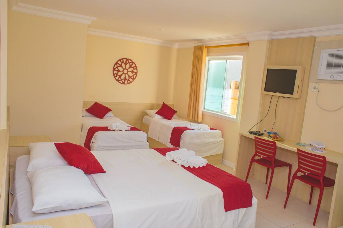 Quarto de hotel com três camas, cada uma com lençóis brancos e almofadas vermelhas, uma escrivaninha com cadeira e uma televisão fixada na parede.