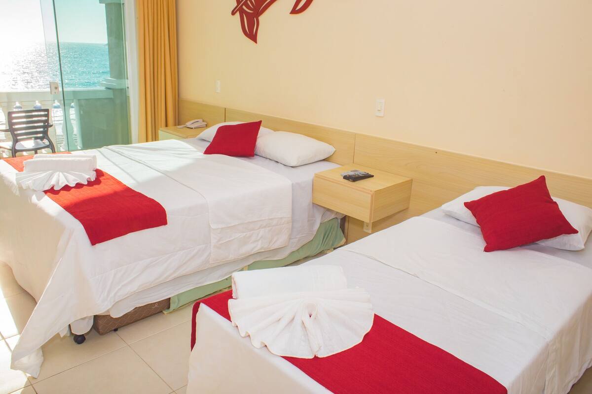 Quarto de hotel com duas camas, cada uma com roupa de cama branca e detalhes vermelhos, uma mesa de cabeceira de madeira com telefone, e vista para o mar através de uma grande janela.