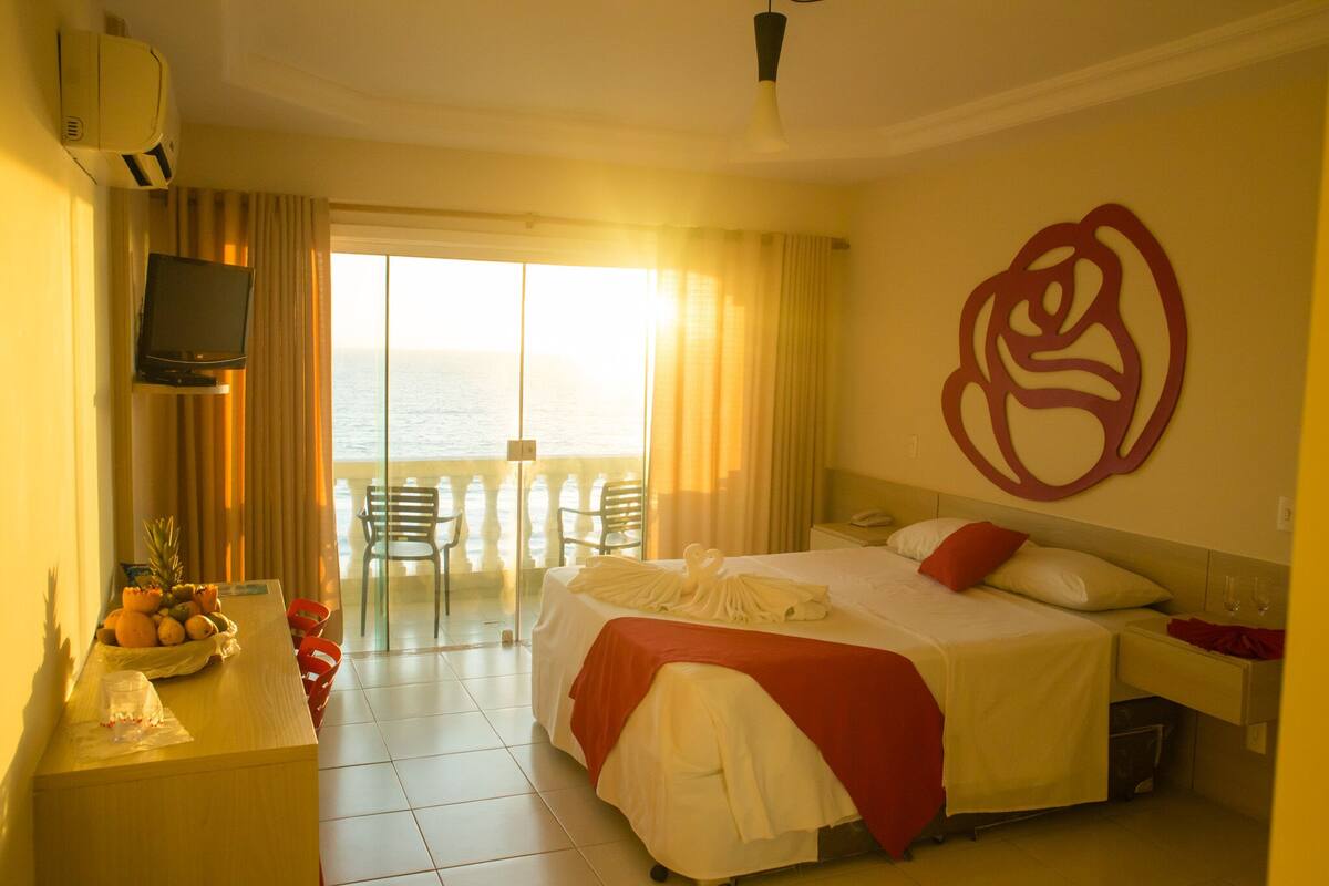 Quarto de hotel com cama, televisão, cesta de frutas e varanda vista para o mar.