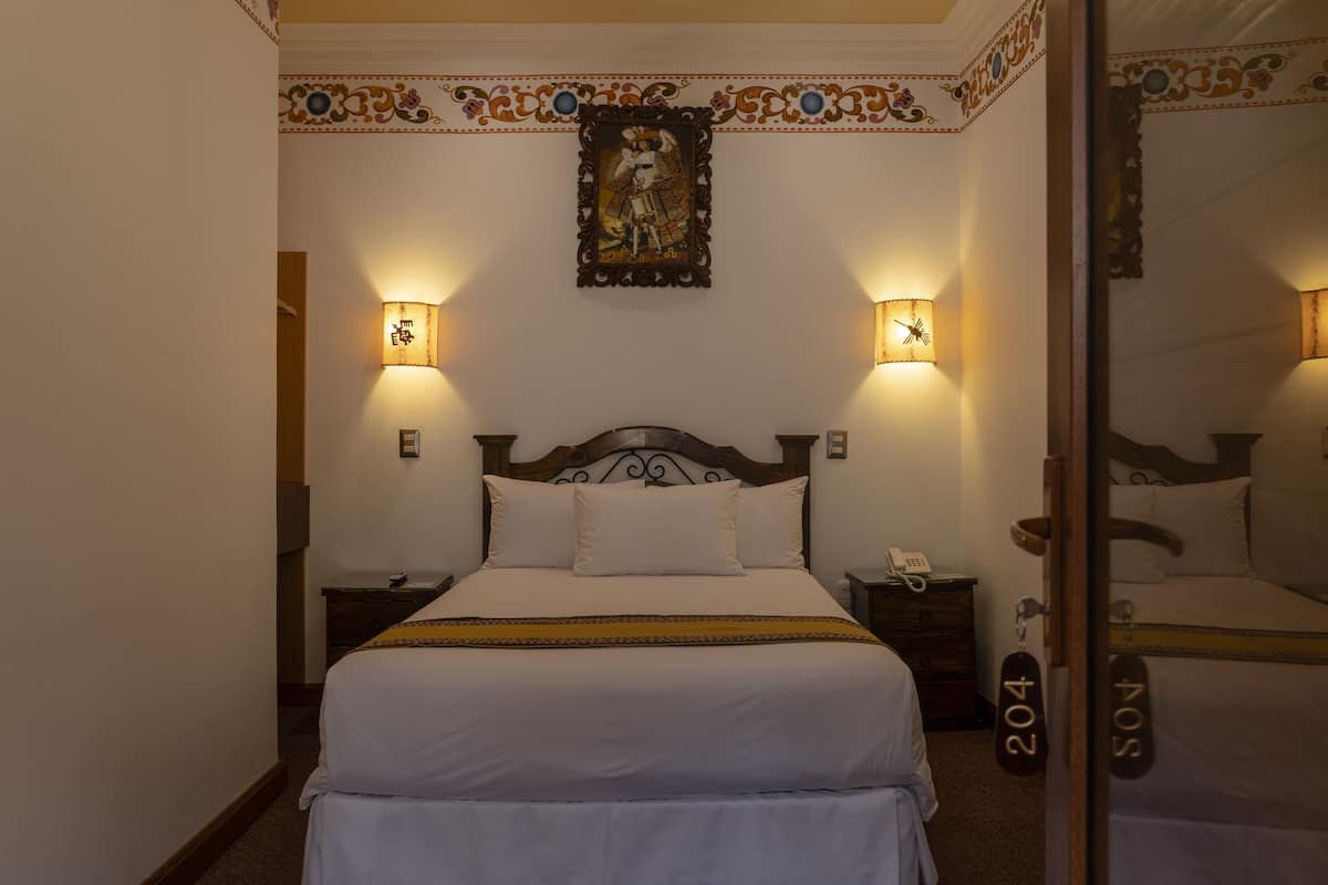 Quarto de hotel com uma cama grande, duas mesas de cabeceira, um criado-mudo, um espelho e um painel decorativo na parede.