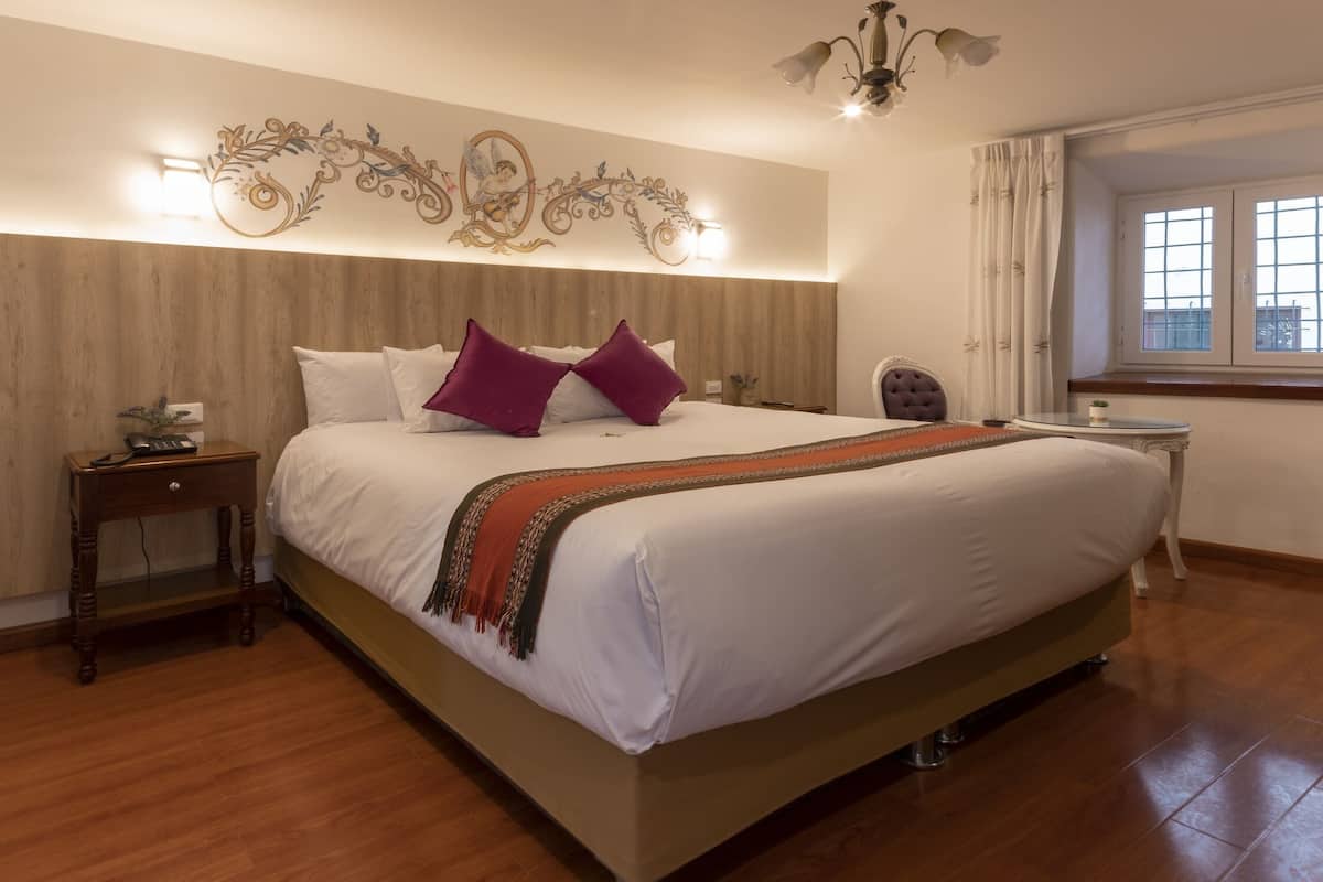 Quarto de hotel com uma cama grande, uma mesinha de cabeceira, uma cadeira, uma pequena mesa e uma janela com cortinas.