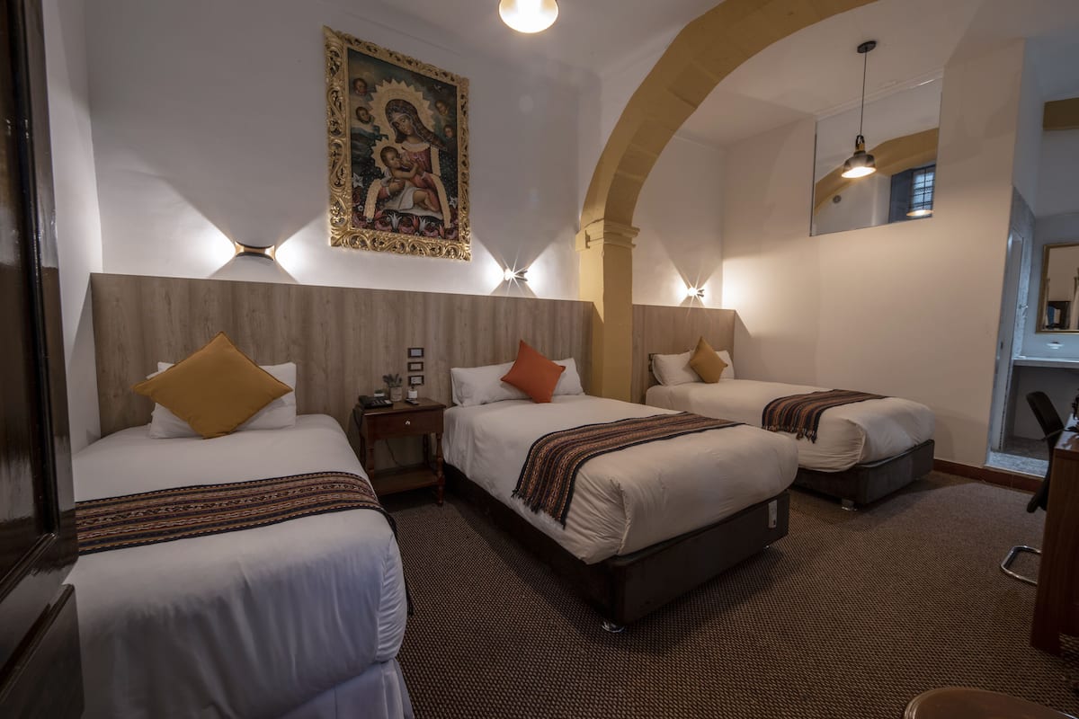 Quarto de hotel com três camas, cabeceira de madeira, pintura religiosa grande e um espelho.