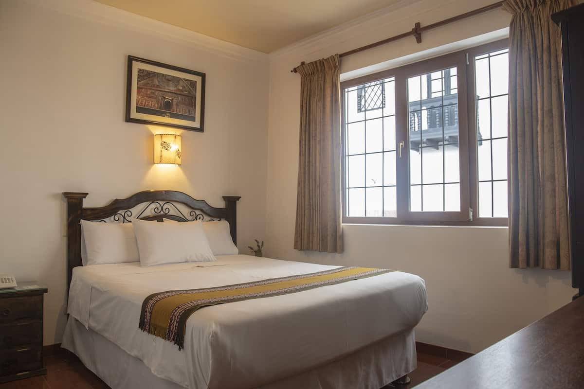 Uma cama bem arrumada com cabeceira, luminária na mesinha de cabeceira, quadro na parede e janela com cortinas.