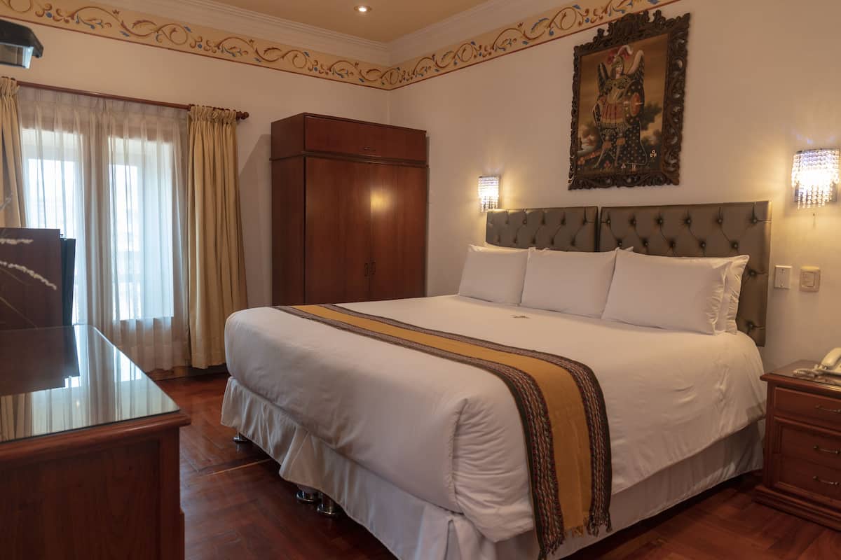 Quarto de hotel com uma cama grande, um guarda-roupa de madeira, uma escrivaninha e um candelabro.