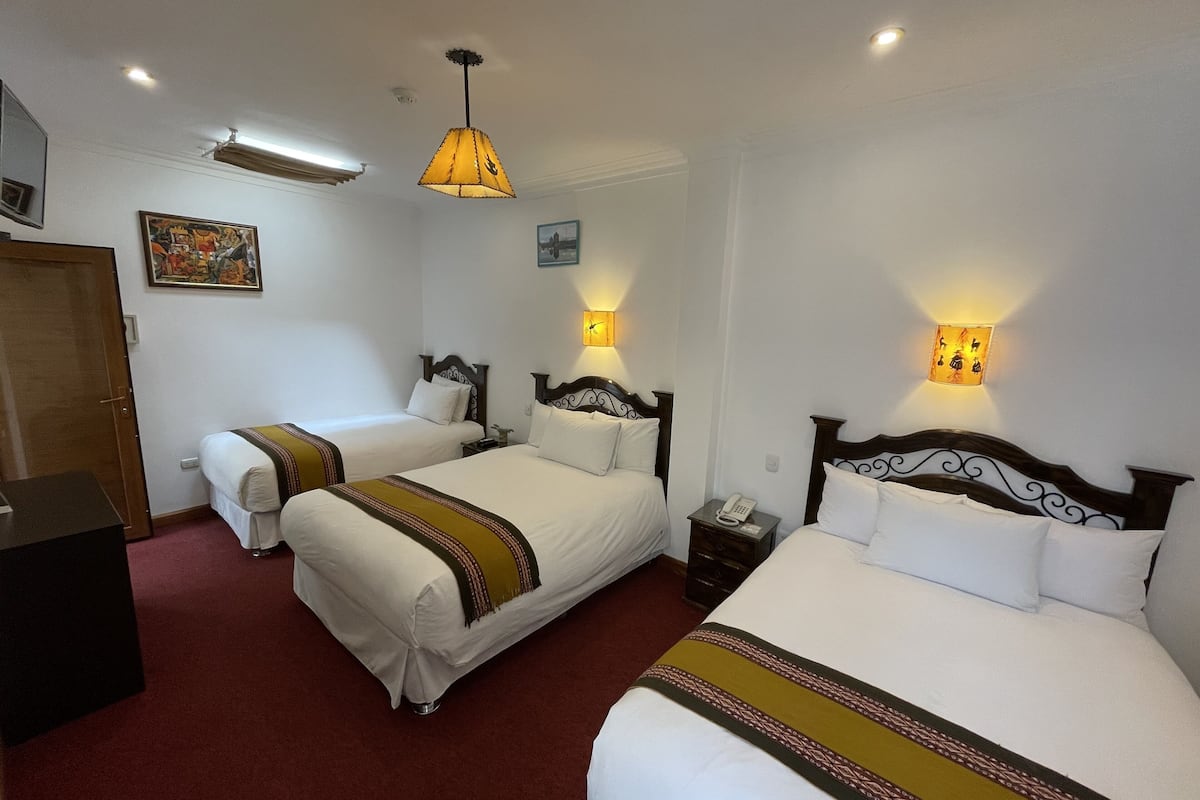 Quarto de hotel com duas camas de solteiro, um guarda-roupa de madeira, uma escrivaninha e uma mesa pequena.