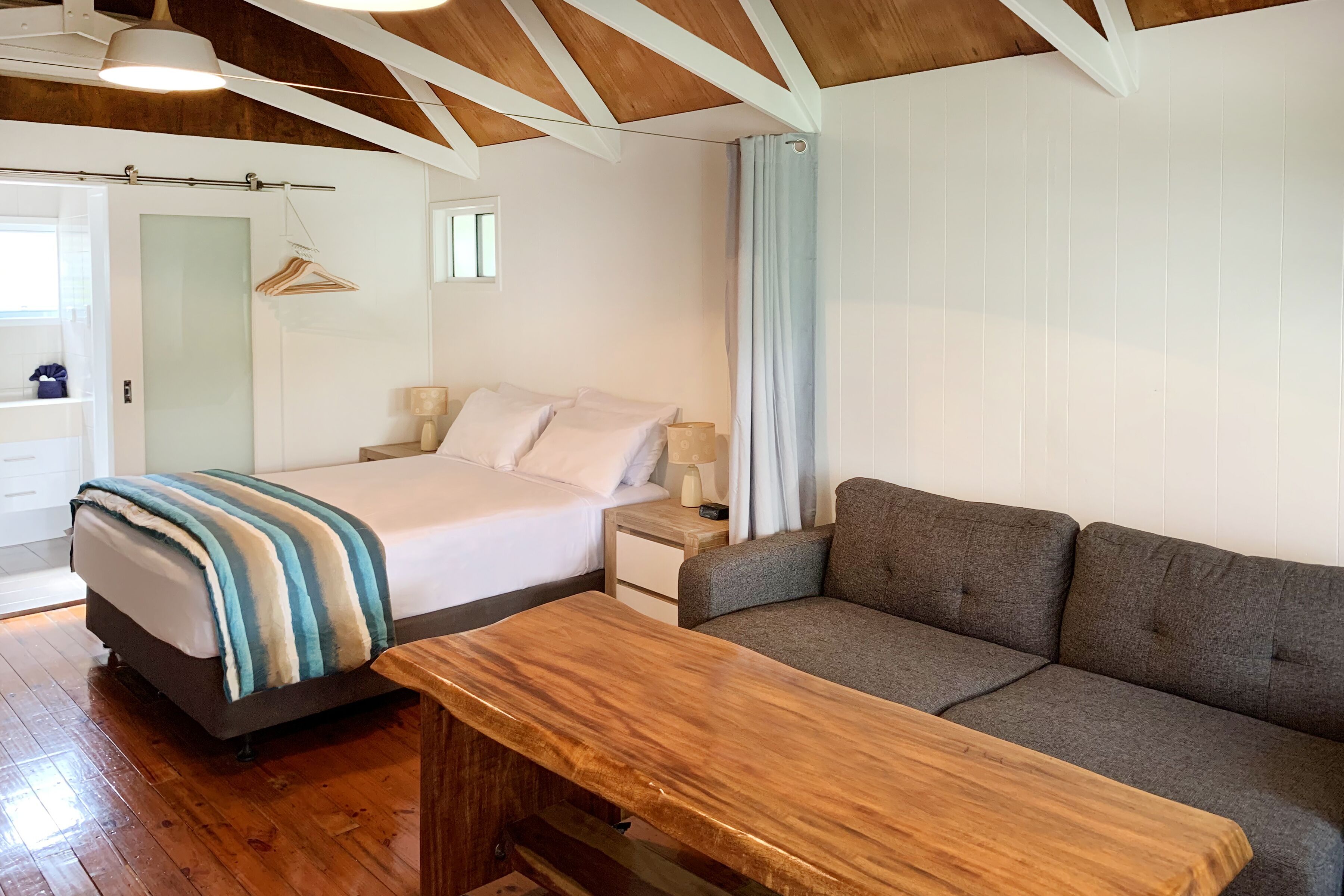Quarto com cama, um banquinho de madeira, um sofá cinza e uma mesa de cabeceira com abajur.