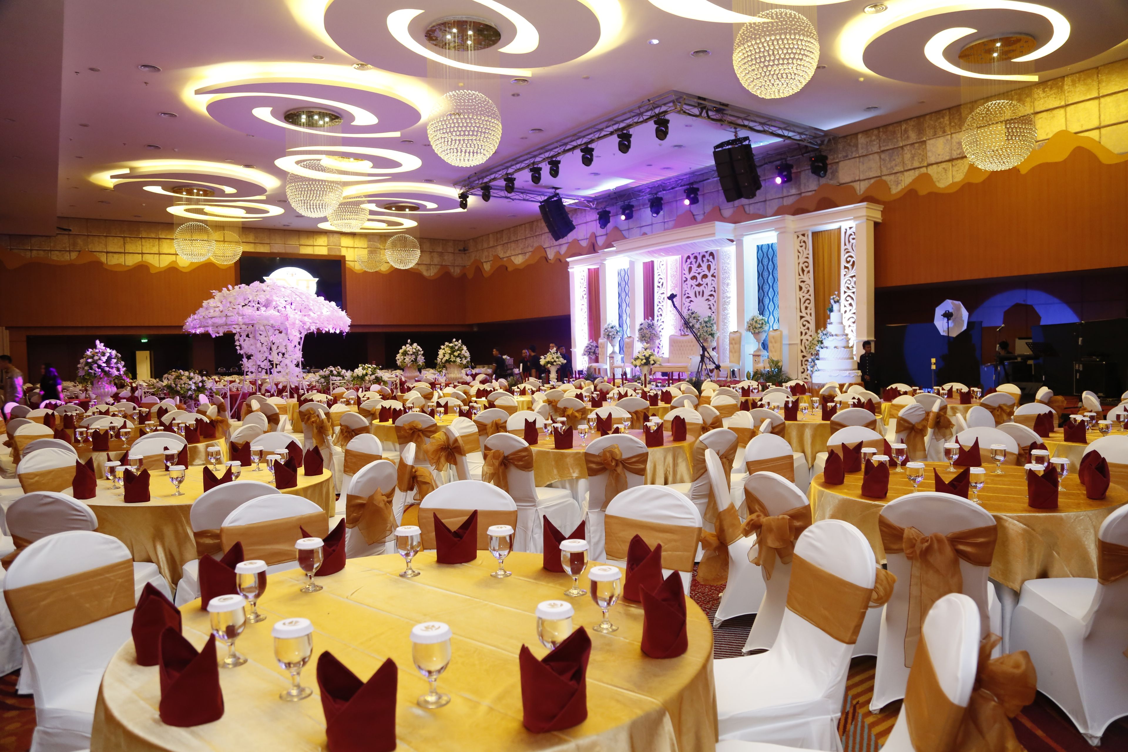 Uma sala de banquetes com mesas redondas, cadeiras brancas e douradas, e um palco com decoração floral.