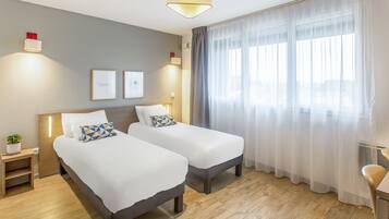 Quarto de hotel com duas camas de solteiro, uma pequena mesa de cabeceira com uma planta, uma escrivaninha e uma cadeira.