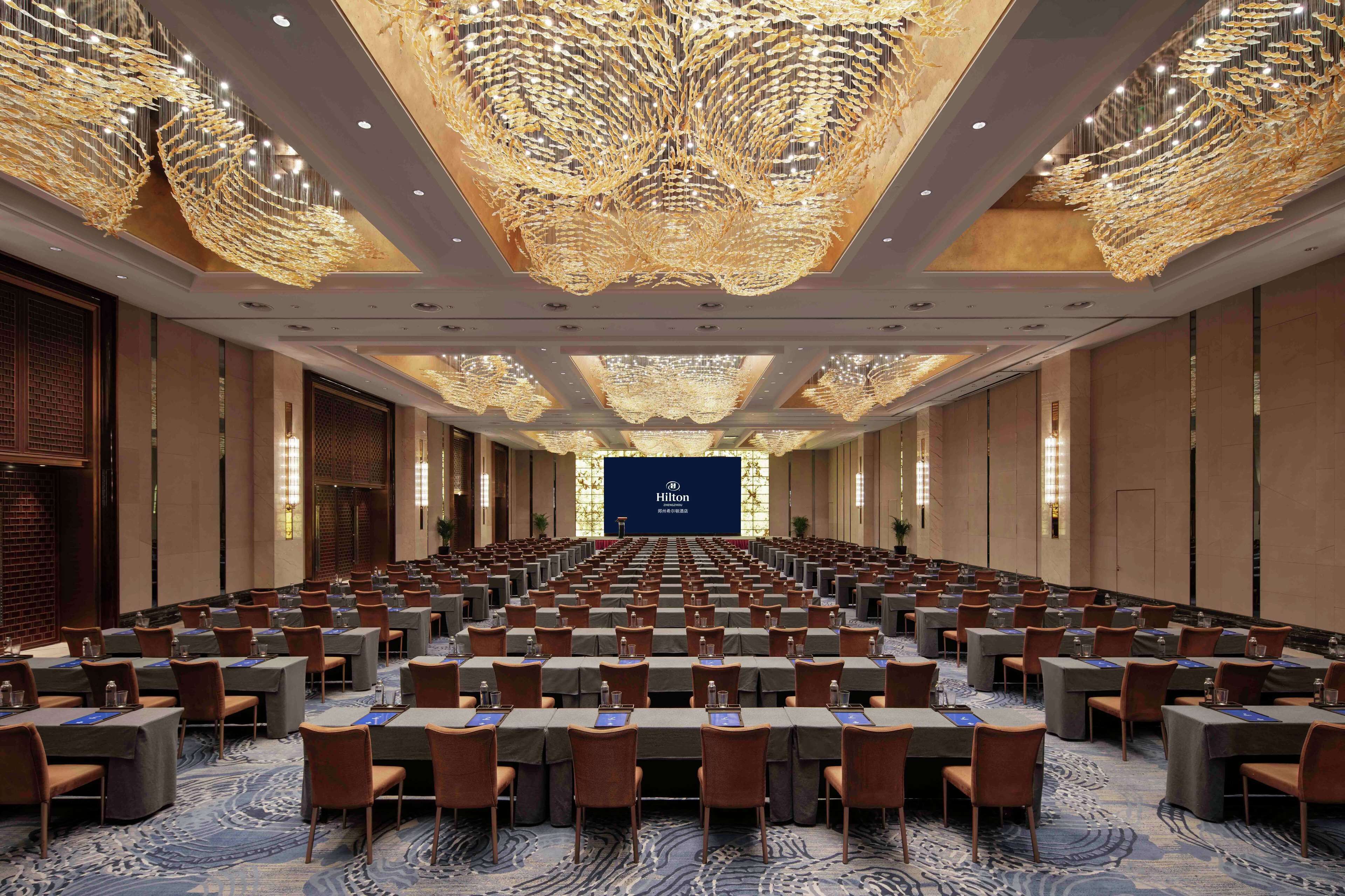 Uma grande sala de conferências com fileiras de mesas e cadeiras, o logotipo da Hilton bem visível na parede de trás e um design de teto ornamentado.