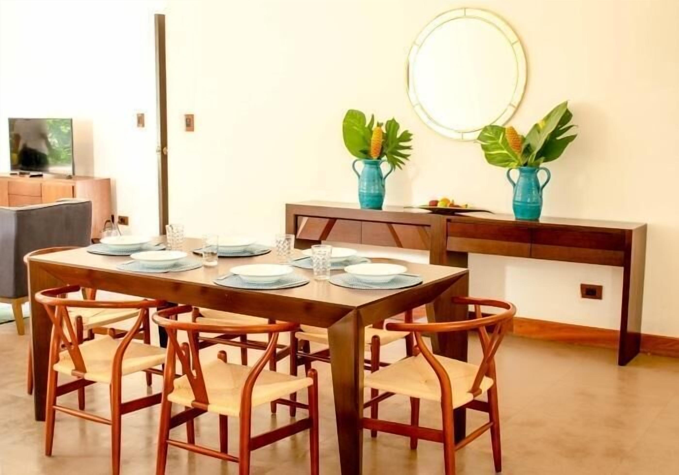 Uma área de refeições com uma mesa de madeira e seis cadeiras, uma TV em um suporte, e dois vasos azuis com plantas em um console de madeira.