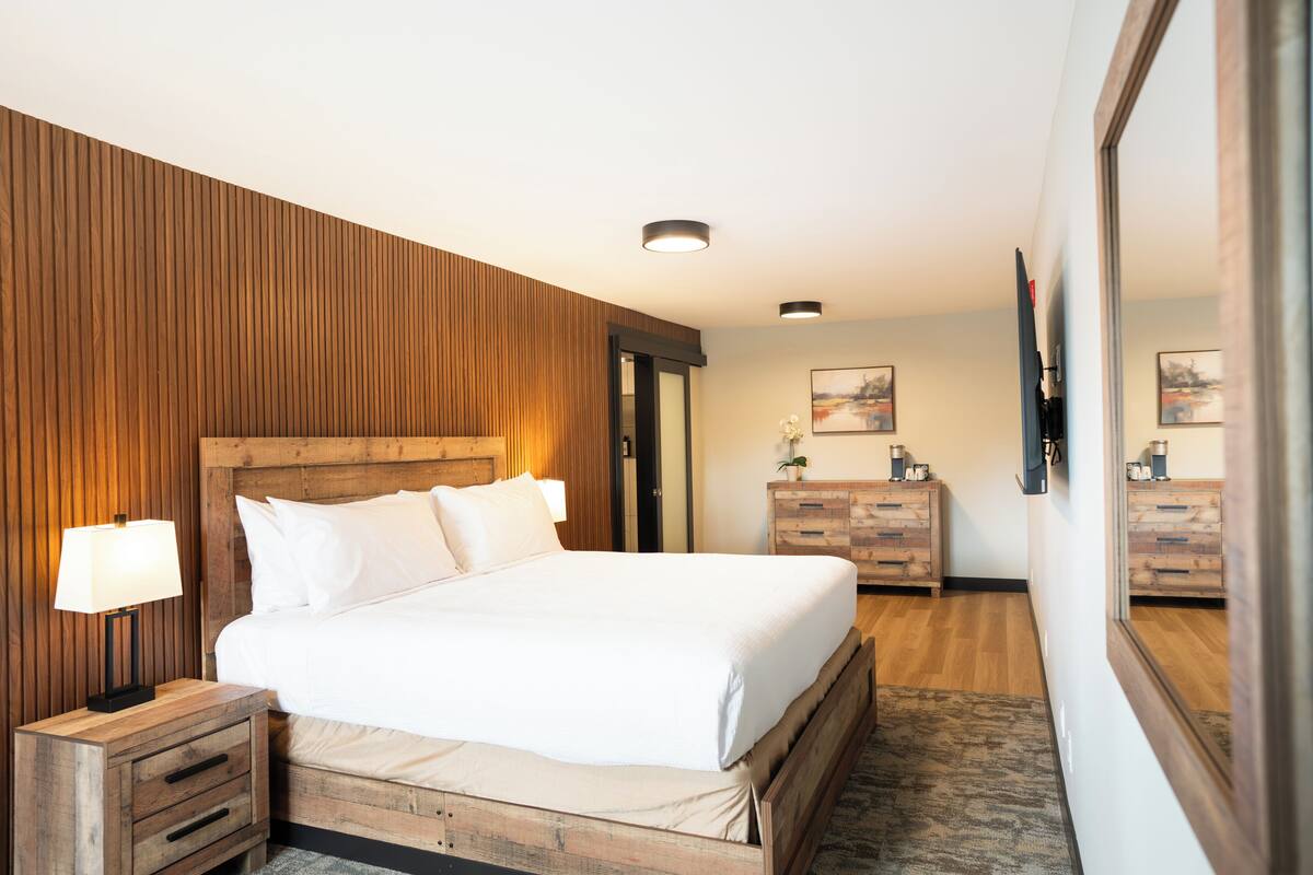 Quarto com cabeceira de madeira, cama com roupa de cama branca, mesa de cabeceira com abajur, penteadeira e espelho.