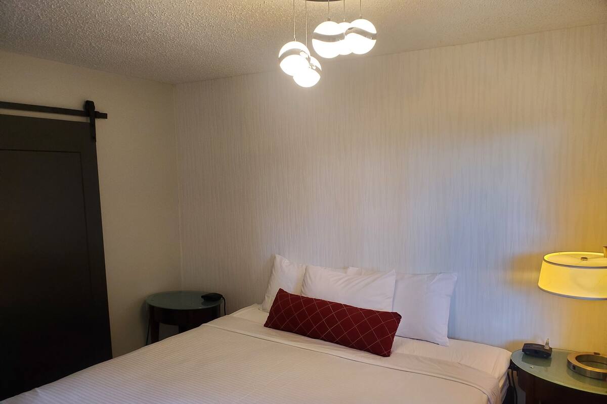 Uma cama bem arrumada com roupa de cama branca e um travesseiro vermelho de destaque, uma mesa de cabeceira com abajur e um lustre no teto.