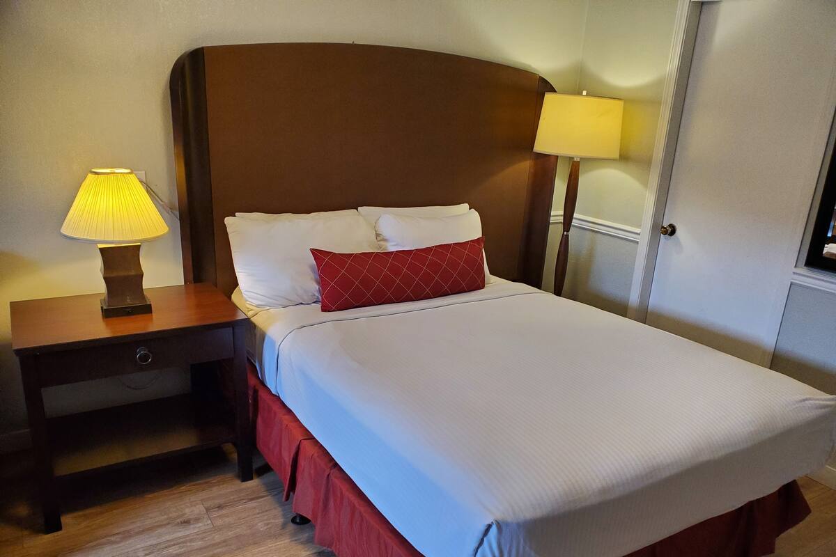 Cama bem arrumada com roupa de cama branca e vermelha, mesa de cabeceira de madeira com abajur e cabeceira de madeira.