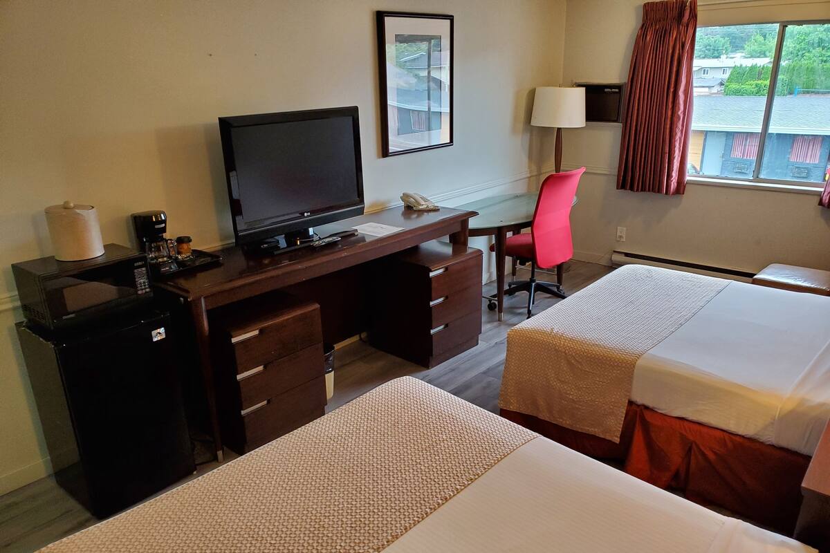 Quarto de hotel com escrivaninha, televisão, cadeira, duas camas e janela com cortinas.