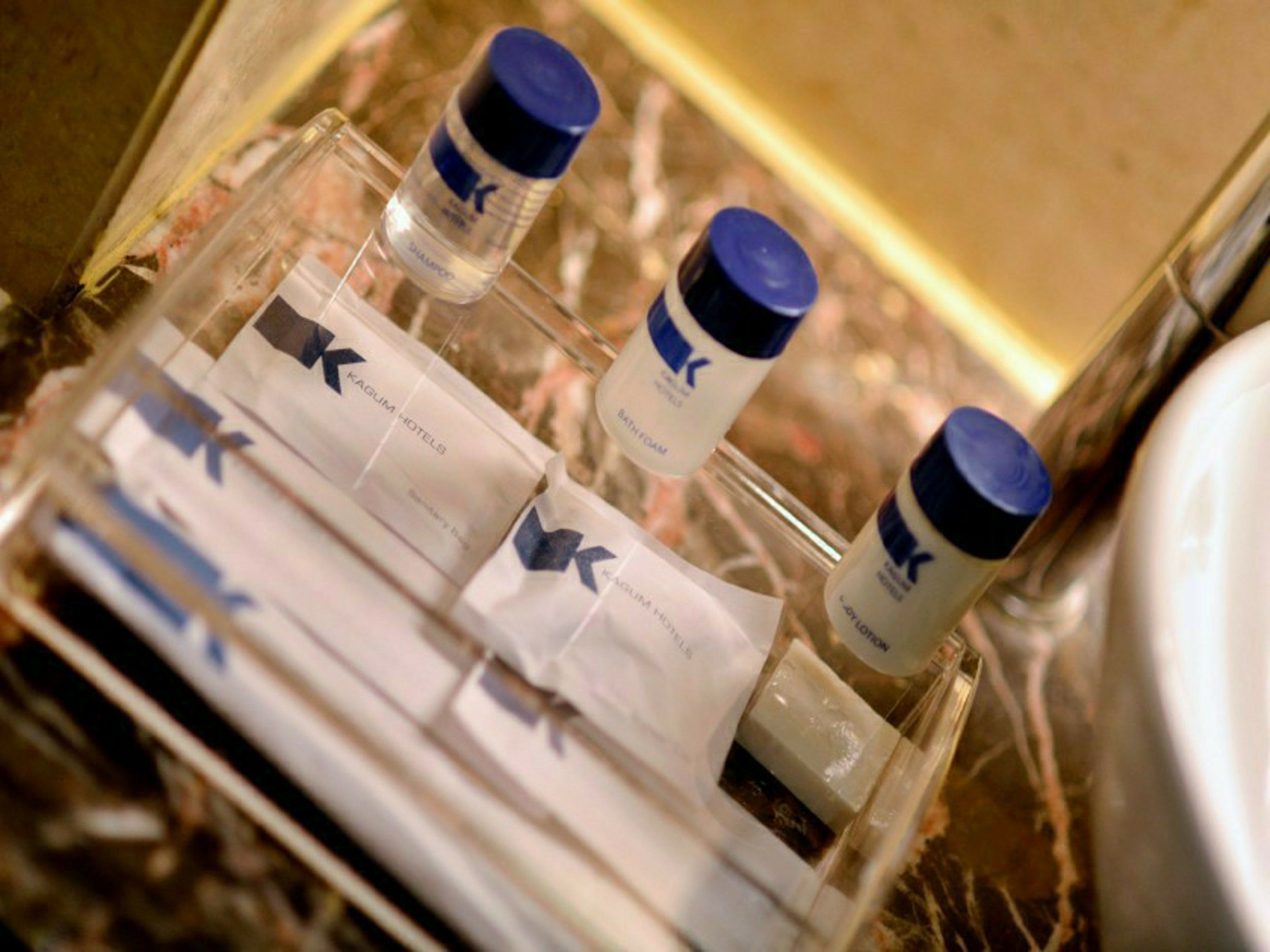 Um conjunto de artigos de higiene pessoal em uma bandeja de plástico transparente com uma etiqueta azul, incluindo um frasco de shampoo e um frasco de loção.