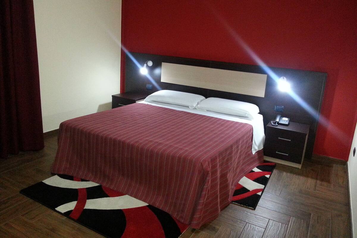 Quarto de hotel com cama, mesa de cabeceira, telefone e parede vermelha.