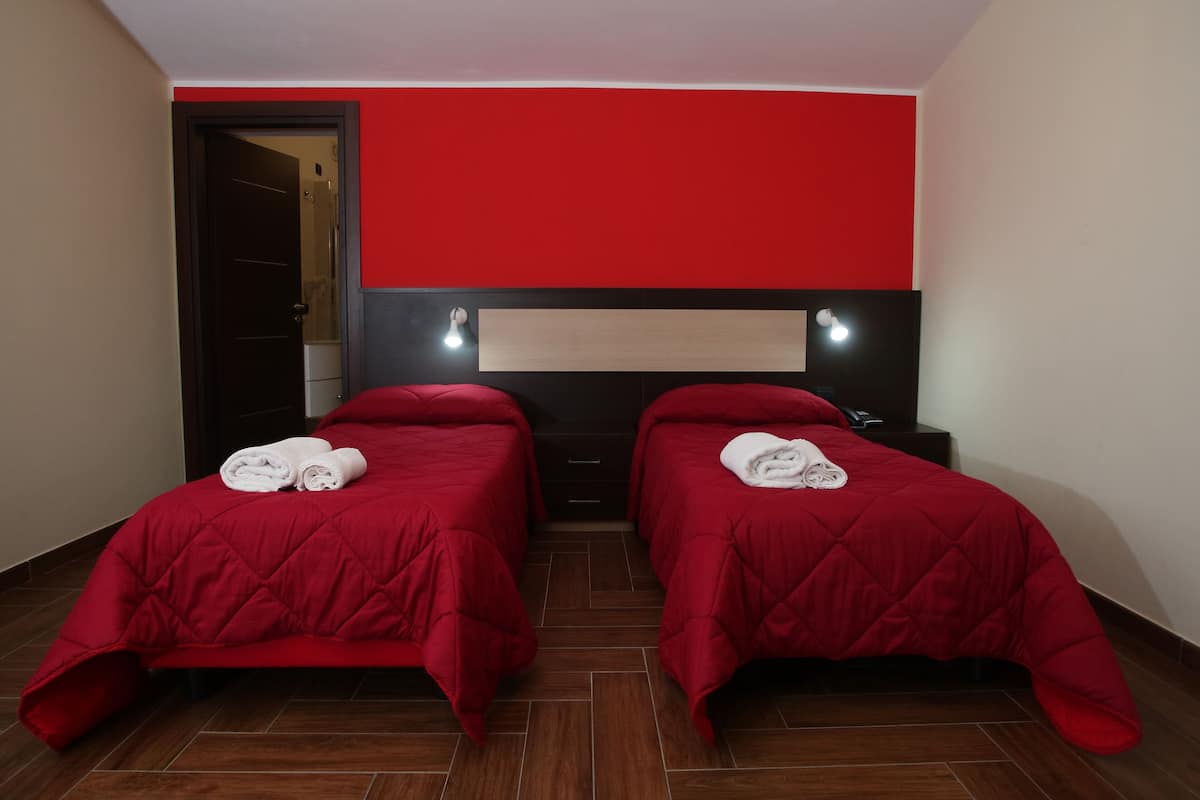 Quarto de hotel com duas camas, roupa de cama vermelha, piso de madeira, parede de destaque vermelha e cabeceira moderna.