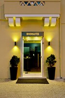 Entrada dos Riversuites, com vista clara do interior através das portas de vidro, um tapete preto e duas plantas em vasos de cada lado.