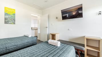 Quarto de hotel com duas camas de solteiro, uma pequena escrivaninha de madeira, televisão fixada na parede e uma janela com persianas.