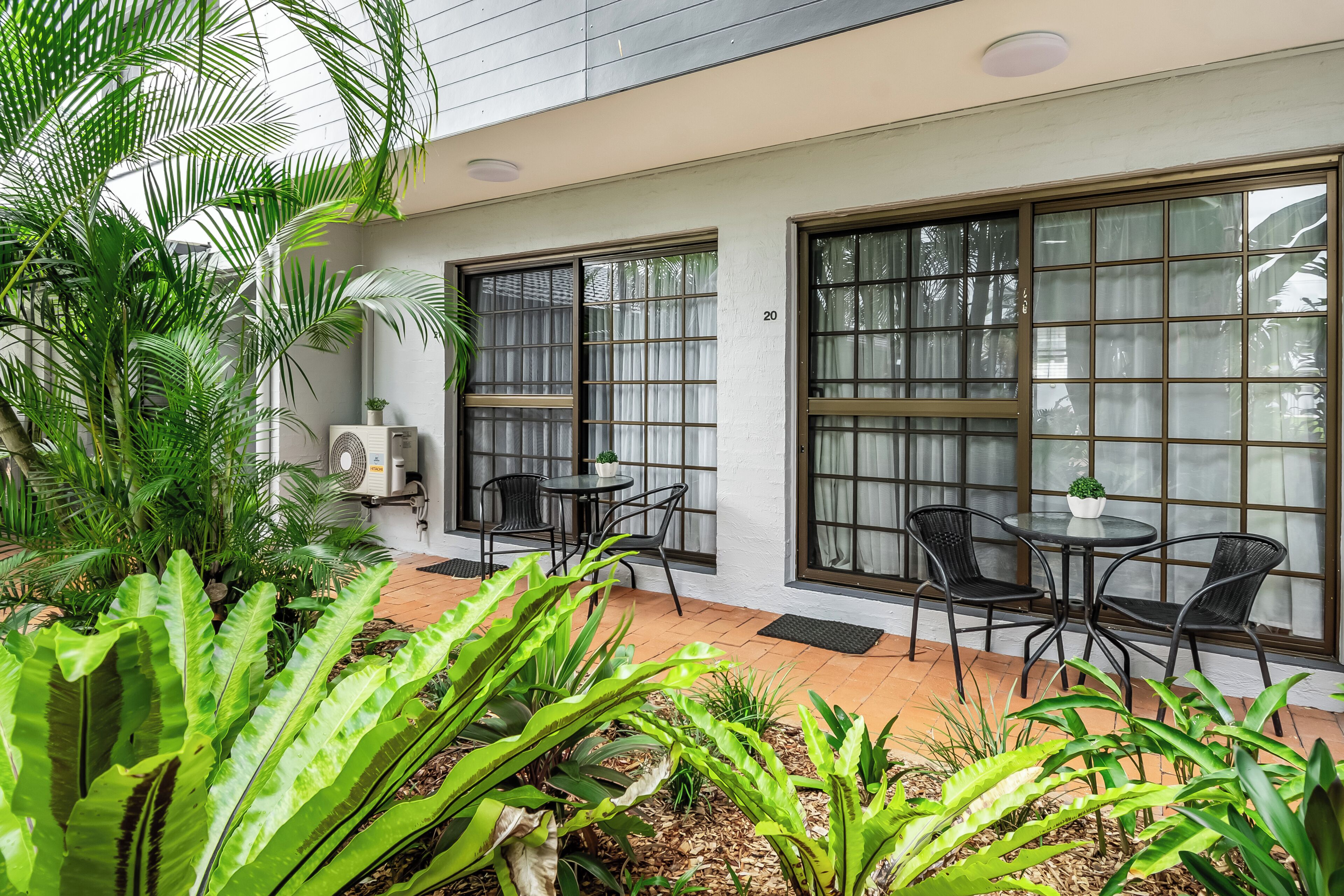 Uma área de pátio com mesa e cadeiras, janelas amplas e vegetação exuberante.