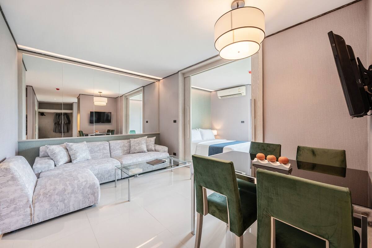 Quarto de hotel moderno com área de estar, mesa de jantar com cadeiras, sofá, televisão e cama.