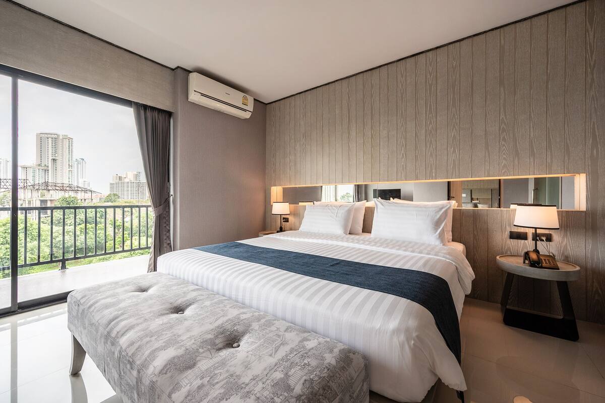 Quarto de hotel moderno com uma cama grande, uma mesa de cabeceira com abajur, uma mesinha redonda com telefone e varanda com vista para a cidade.