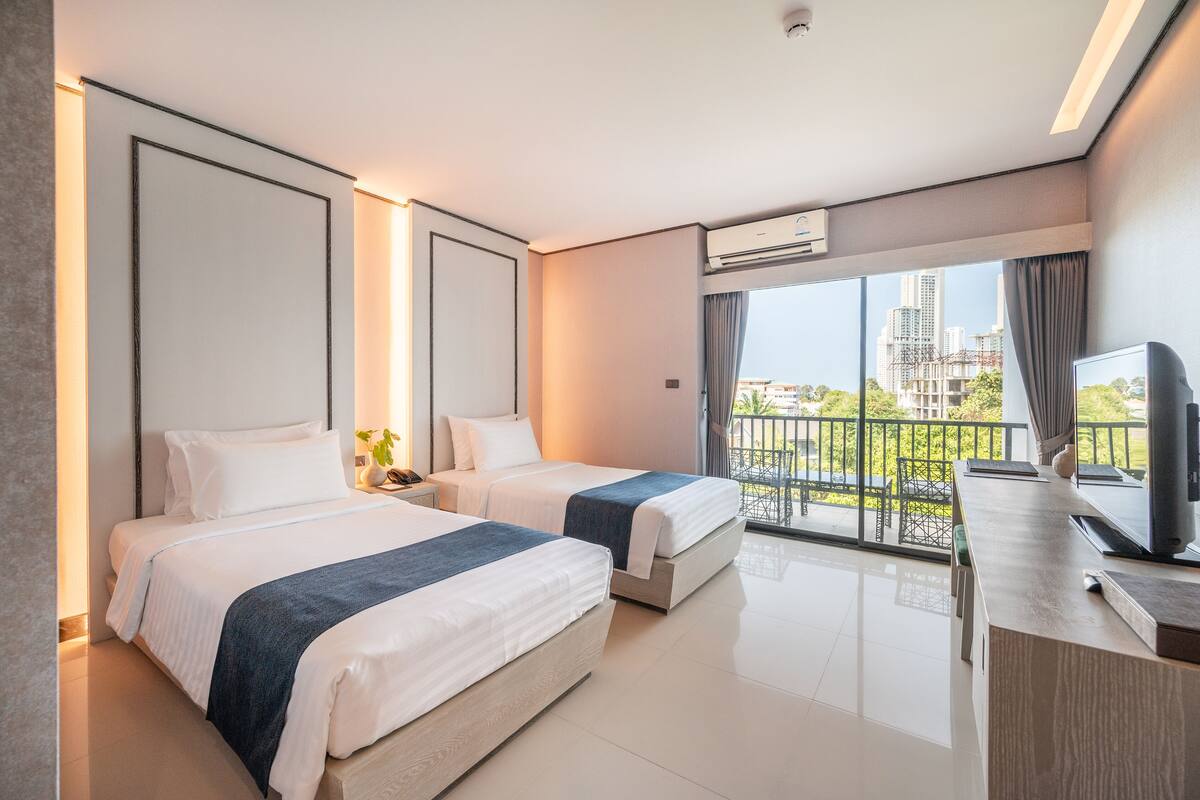 Quarto de hotel com duas camas, uma escrivaninha, televisão e varanda com vista para a cidade.