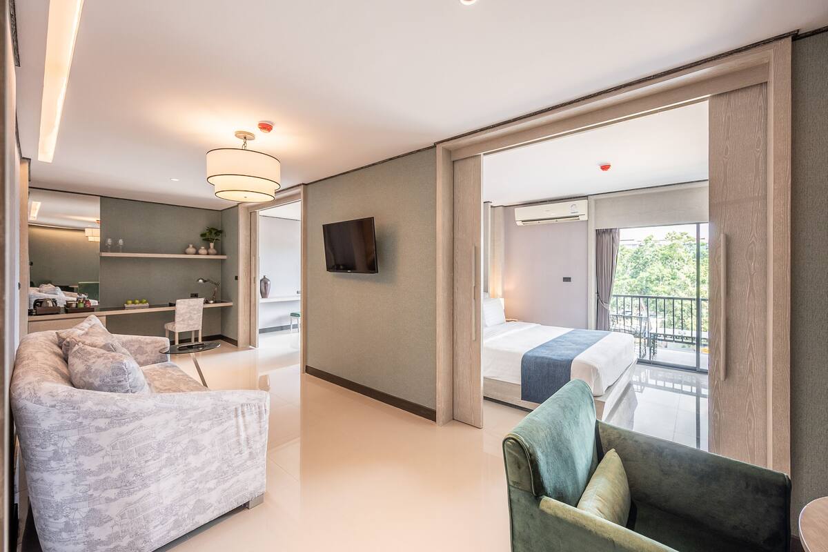 Quarto de hotel moderno com sofá, poltrona verde, cama, televisão e varanda com vista.