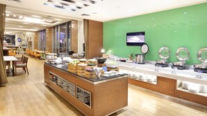 Uma área de buffet com parede verde, uma tela de televisão e diversas opções de comida.