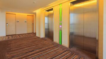 Corredor de hotel com piso revestido de carpete, dois elevadores e portas numeradas 04 e 05.